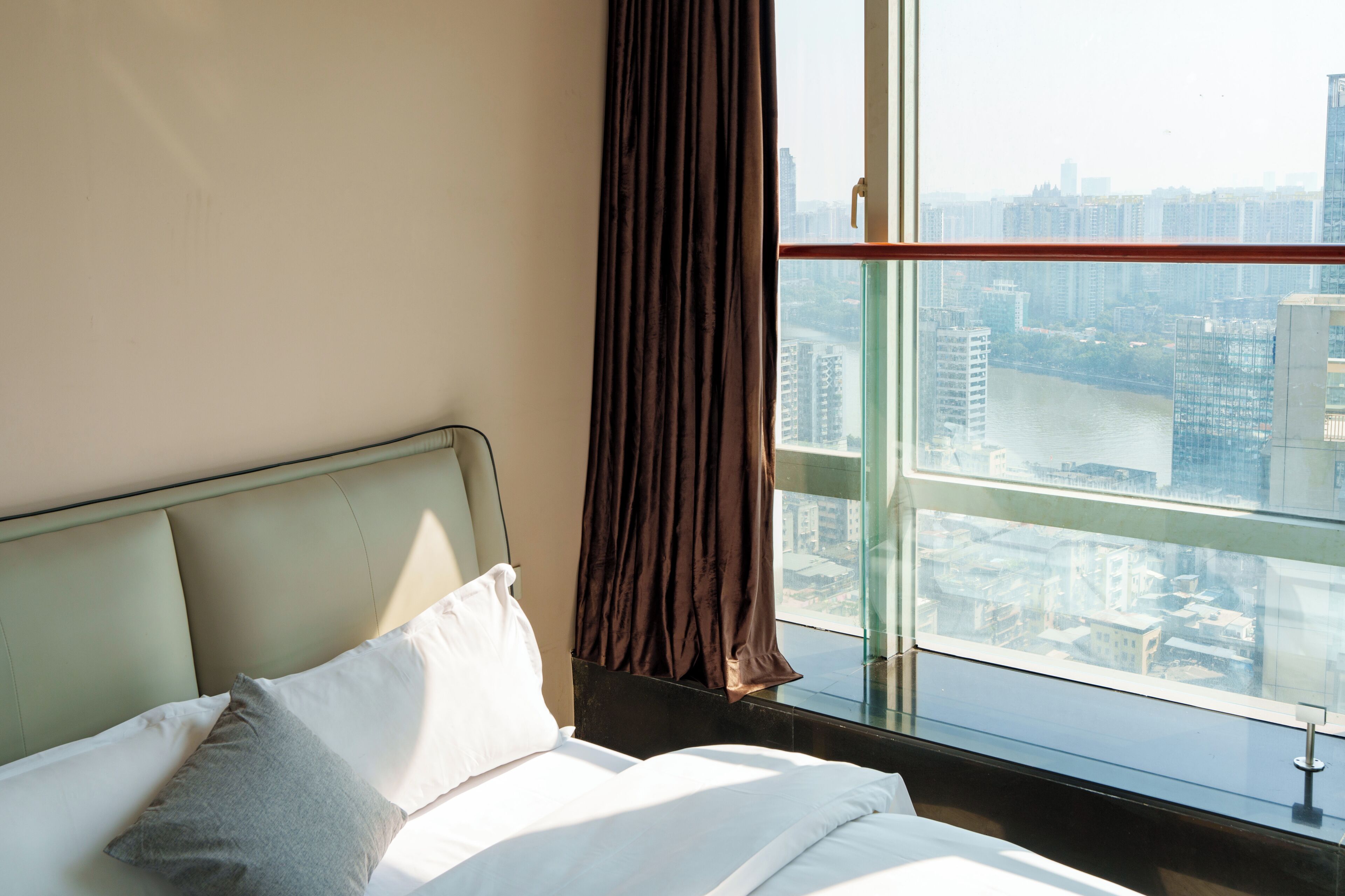 Quarto de hotel com uma grande janela, cama com roupa de cama branca e cinza, e vista para a paisagem urbana.