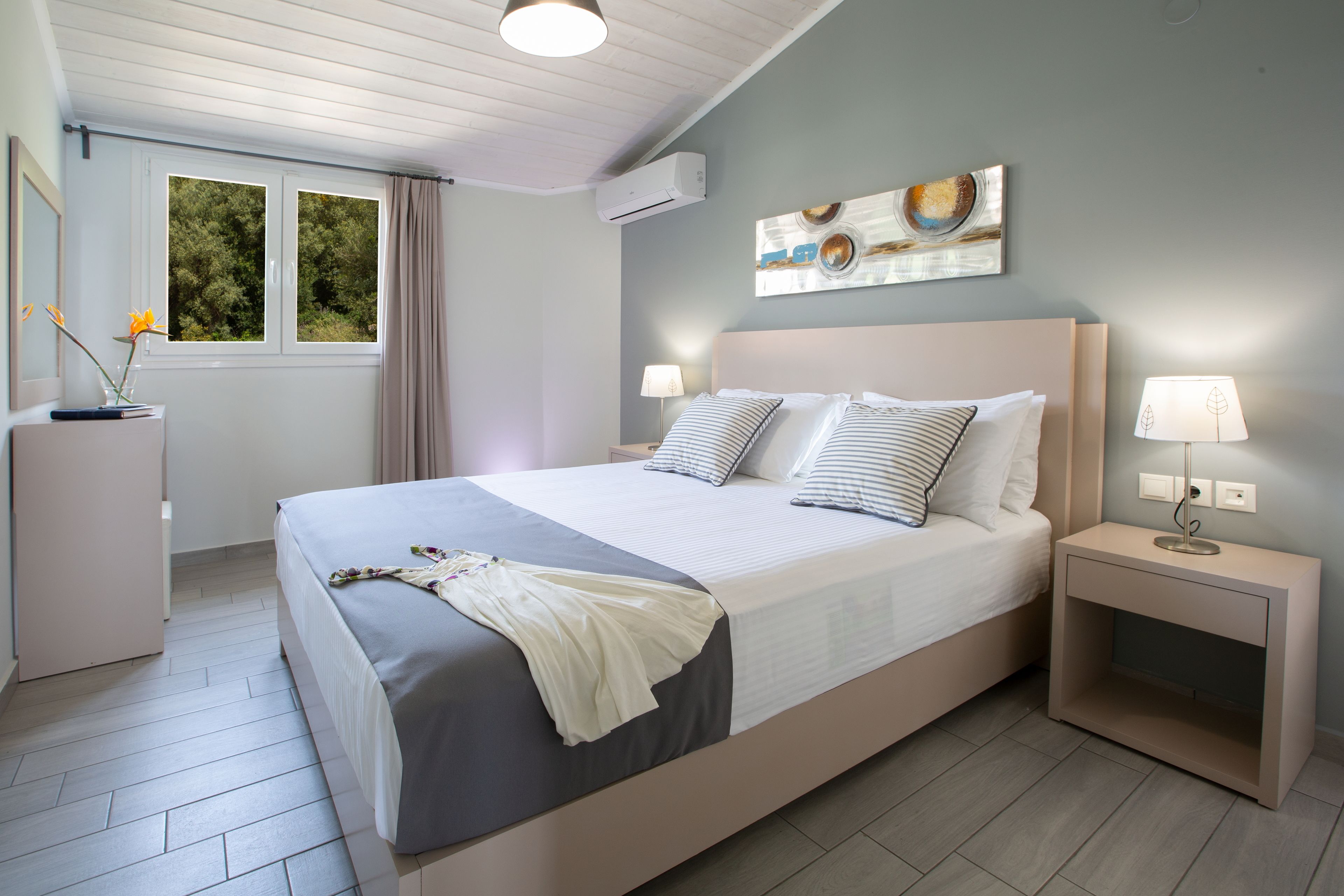 Quarto moderno com uma cama grande, mesas de cabeceira com luminárias, uma janela com cortinas e um ar condicionado fixado na parede.