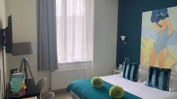 Quarto de hotel com cama, escrivaninha, abajur, radiador e uma grande pintura abstrata na parede.