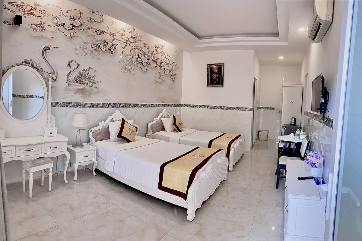 Quarto de hotel com duas camas, uma toucador com espelho, uma secretária e uma parede com arte de flores e pássaros.