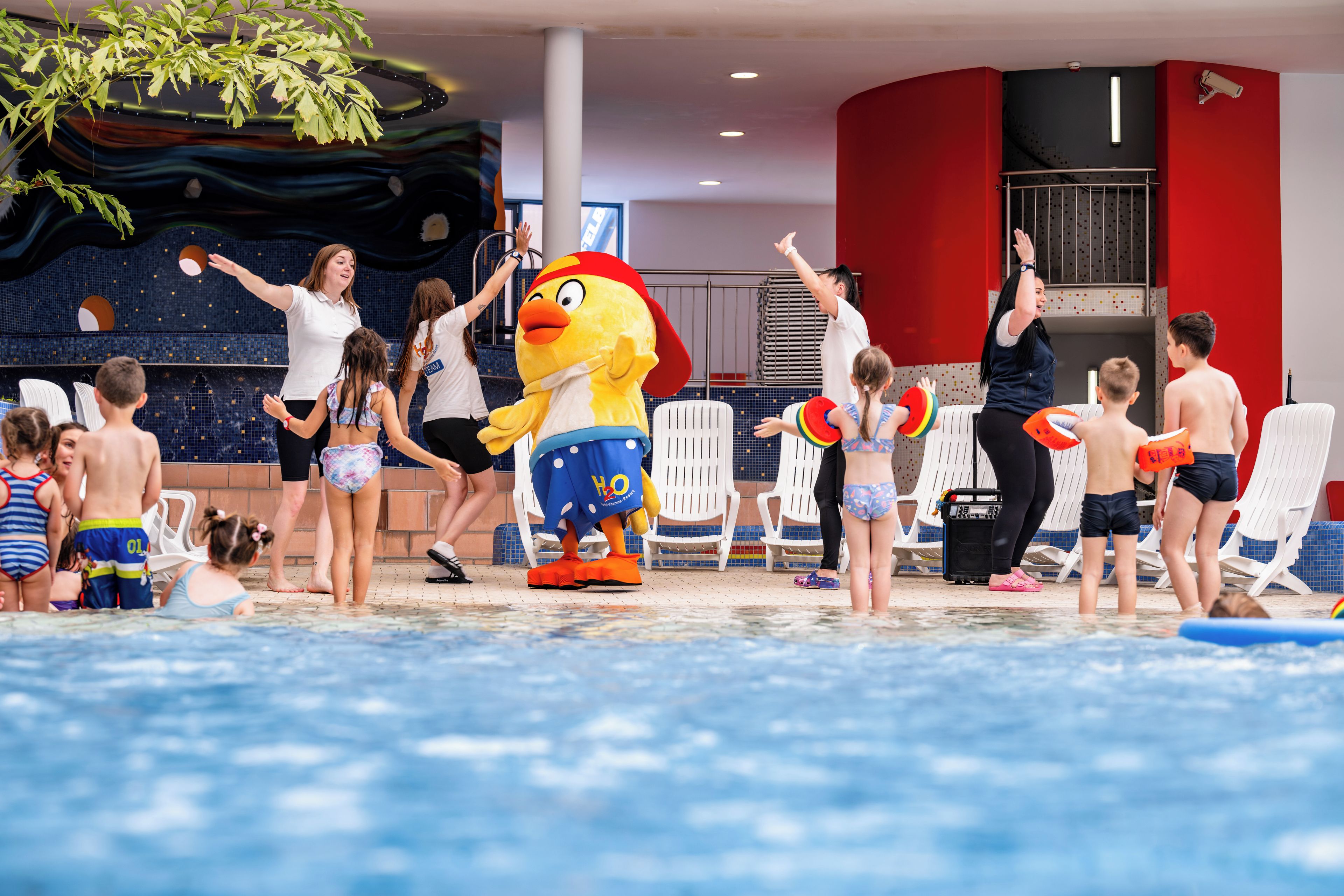 Área de piscina com um mascote, pessoas e espreguiçadeiras.
