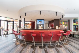 Uma área de bar moderna com um balcão curvo, vários banquetas vermelhas e uma tela de televisão sobre o balcão.