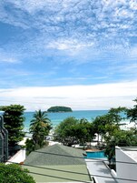 Vista de uma praia com uma ilha pequena, céu claro e vegetação exuberante.