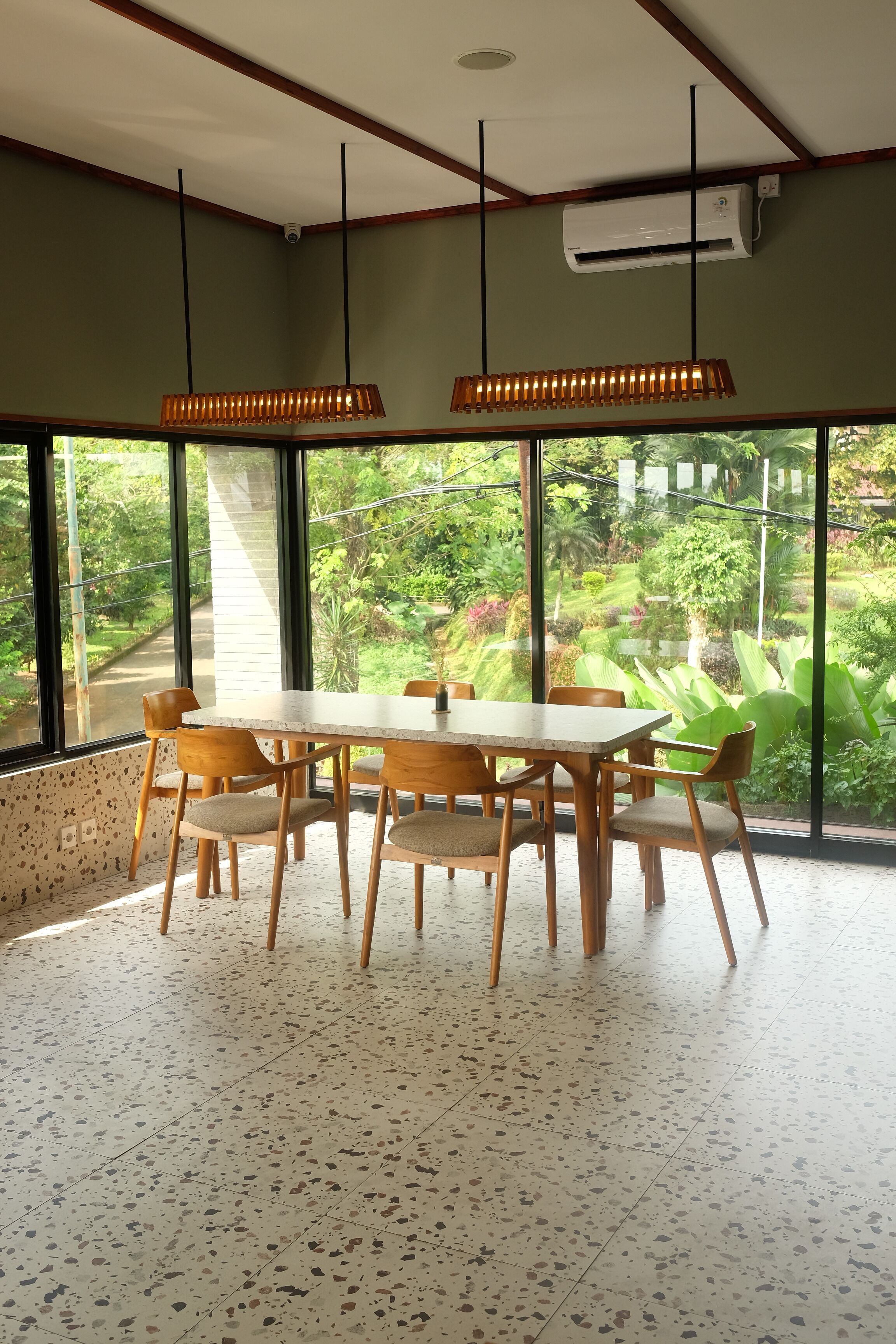 Uma sala de jantar com uma mesa grande e cadeiras, janelas amplas com vista para áreas verdes, e um aparelho de ar condicionado na parede.