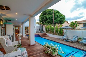 Área de piscina com deck de madeira, cadeiras de descanso e uma grande árvore ao fundo.