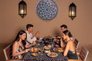 Uma mesa de jantar com toalha de mesa estampada, rodeada por cadeiras, e um pendente decorativo acima.