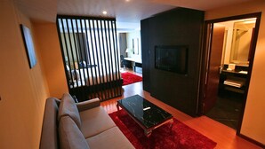Uma sala de estar moderna com sofá, mesa de centro e televisão. Há um tapete vermelho no chão e um painel com persianas verticais.