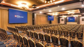 Uma sala de conferências com fileiras de cadeiras, uma tela grande exibindo o logotipo do Hilton Garden Inn e um lustre.