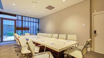 Uma sala de conferências com uma mesa comprida, cadeiras brancas e uma janela grande com persianas.