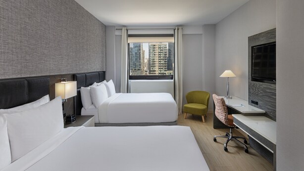 Quarto de hotel com duas camas, uma escrivaninha, uma cadeira, uma TV e uma janela com vista para a cidade.