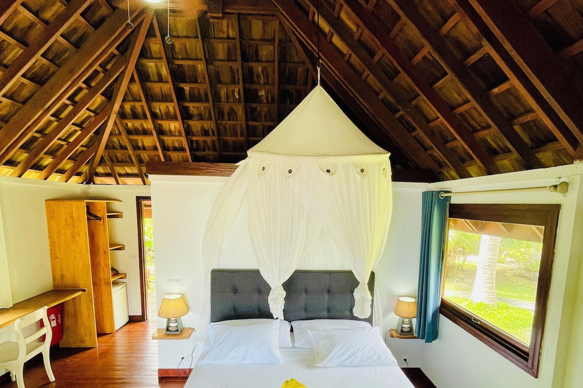 Quarto com cama de dossel, piso de madeira, ventoinha no teto e janela com vista para o verde.