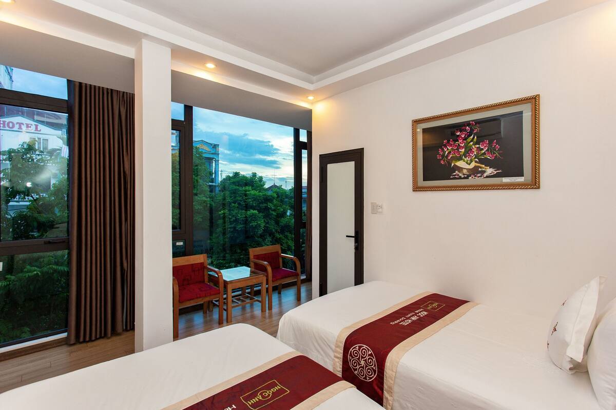 Quarto de hotel com duas camas, área de refeições com mesa e cadeiras, uma janela grande com cortinas e um quadro na parede.