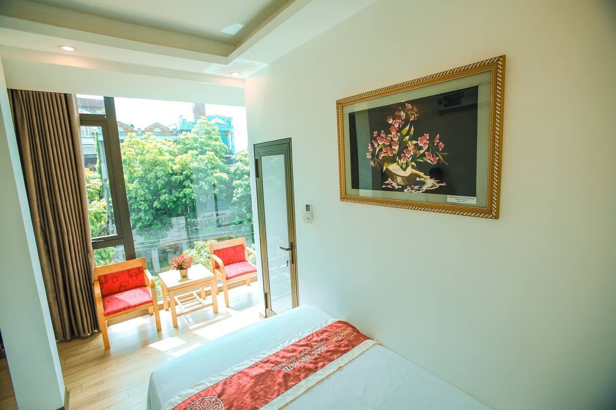 Quarto de hotel com uma cama, uma cadeira vermelha, uma mesa com um vaso de flores e uma grande janela com vista para vegetação e edifícios.