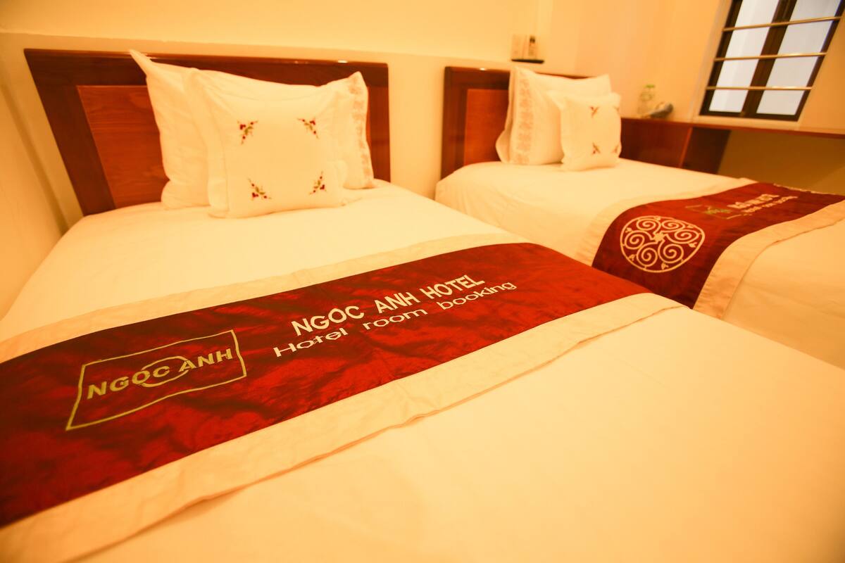 Quarto de hotel com duas camas, cada uma com roupa de cama branca e almofadas decorativas. Há um espalha de cama vermelho com o logotipo e o nome do hotel.