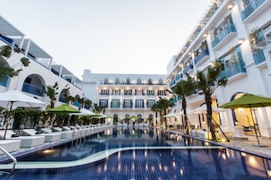Uma área de piscina de hotel com espreguiçadeiras e guarda-sóis, rodeada por edifícios brancos com varandas azuis.