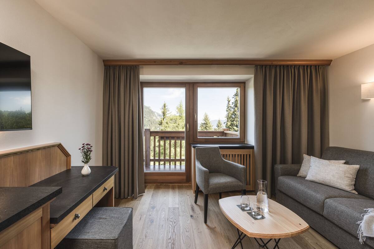 Quarto de hotel moderno com uma secretária de madeira, um sofá cinzento, uma mesinha e uma varanda com cortinas.
