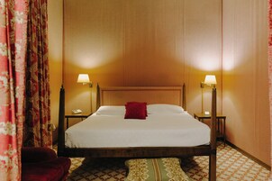 Quarto de hotel com uma cama grande, duas luminárias de cabeceira, um travesseiro vermelho e cortinas estampadas.