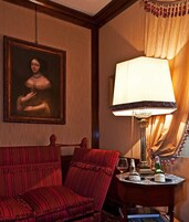 Um quarto com um sofá vermelho estofado, um retrato emoldurado na parede, uma mesa com abajur e garrafas, e uma cortina com franjas.