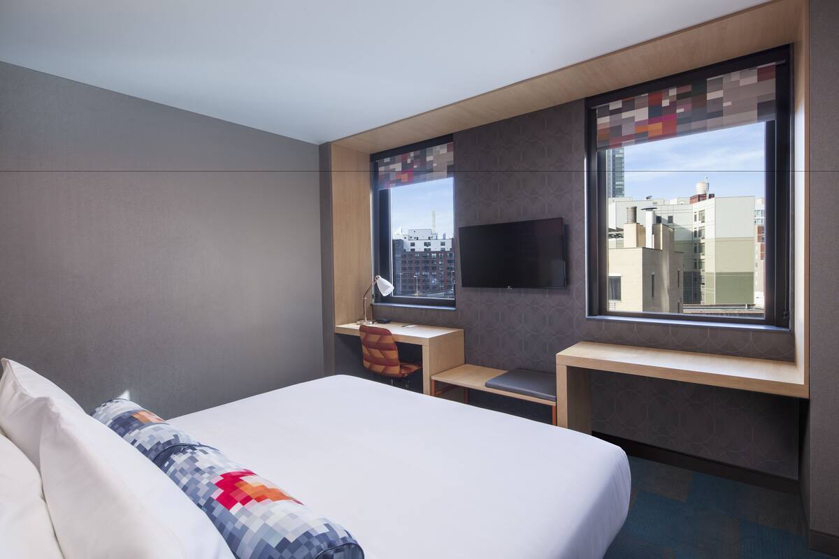 Quarto de hotel com cama, escrivaninha, cadeira, TV e amplas janelas com vista para a cidade.