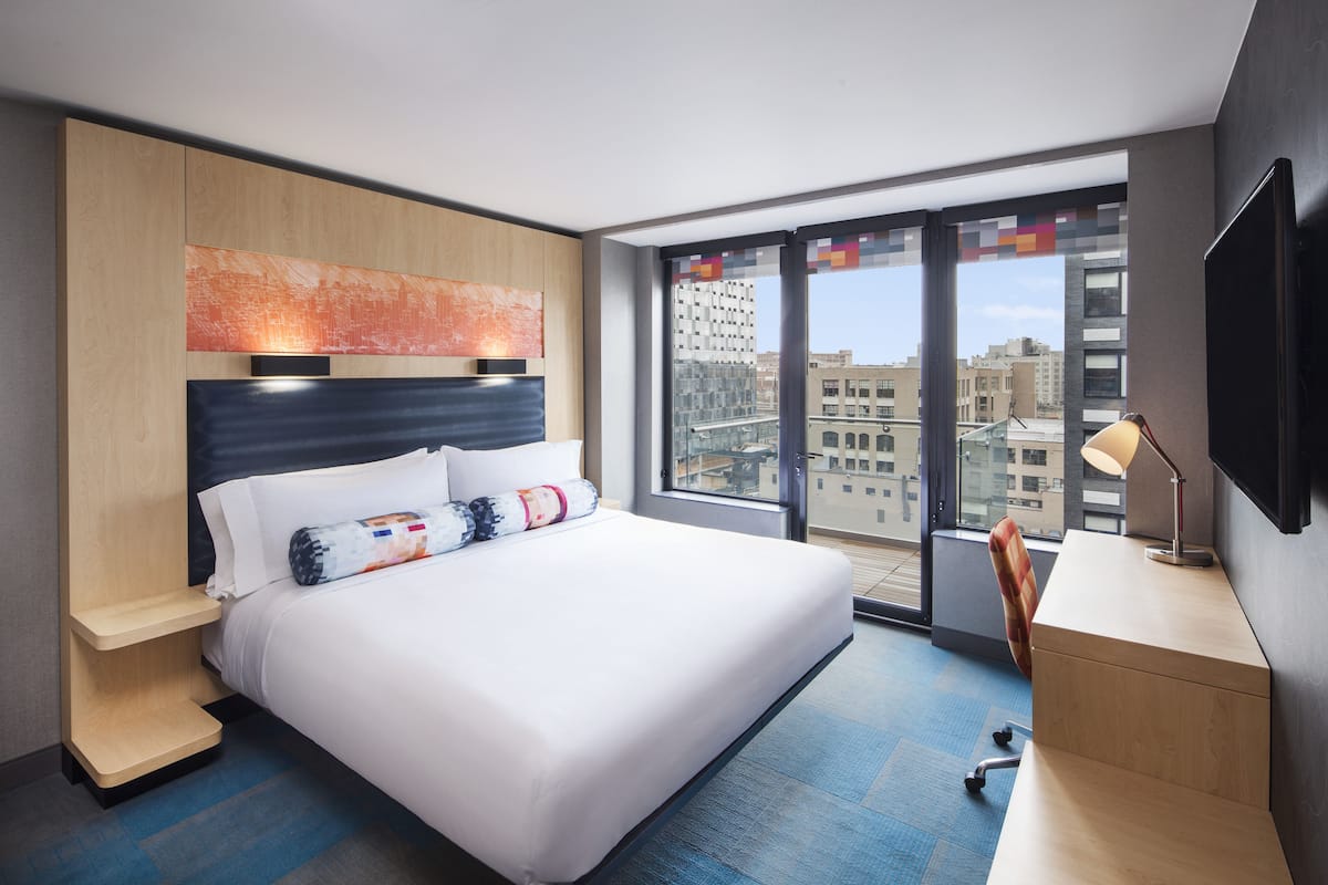 Quarto de hotel moderno com uma cama grande, uma escrivaninha e vista da cidade através de amplas janelas.