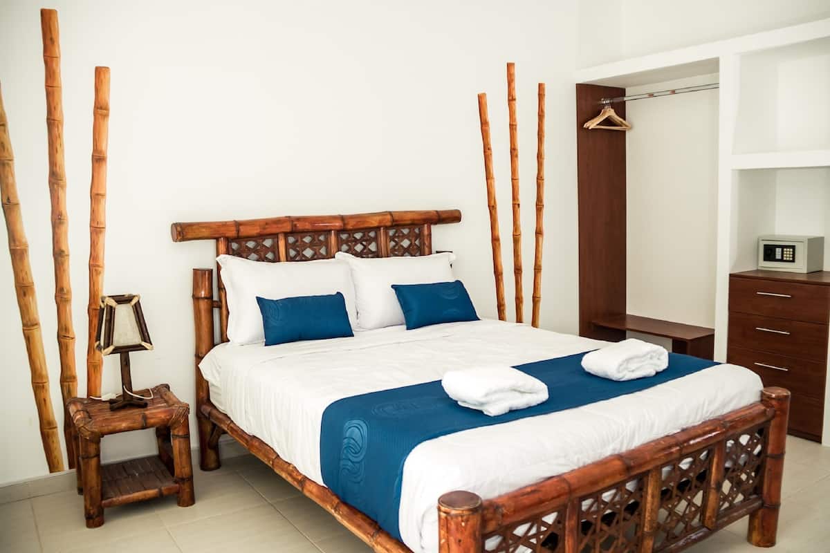 Uma cama de madeira com colcha azul e branca, uma mesa de cabeceira com abajur e um guarda-roupa de madeira.