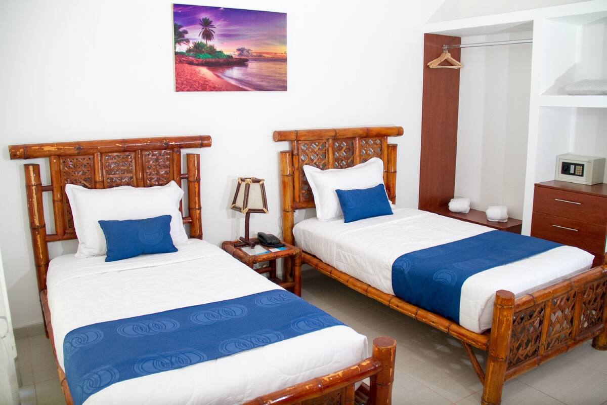 Duas camas de madeira com roupa de cama azul e branca, uma mesa de cabeceira com abajur, um guarda-roupa de madeira e uma pintura de pôr do sol na parede.