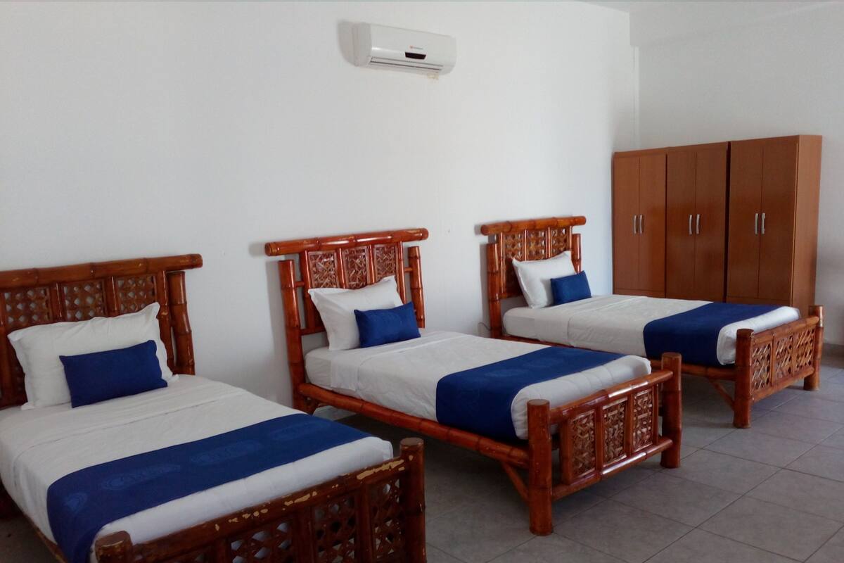Quarto de hotel com três camas de madeira, cada uma com colchão branco e colcha azul. Possui um guarda-roupa de madeira e um aparelho de ar condicionado fixado na parede.