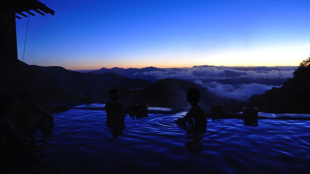 Uma nascente termal tranquila, com duas pessoas apreciando a vista de uma cordilheira ao pôr do sol.