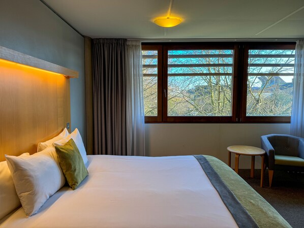 Quarto de hotel com uma cama grande, uma mesinha redonda pequena, uma cadeira e uma janela com vista para árvores e edifícios.