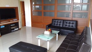 Uma sala de estar com um sofá de couro preto, uma mesa de centro de vidro com um bolo, uma televisão e uma porta de madeira com painéis de vidro jateado.