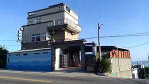 Um edifício com vários andares, com um mural azul na fachada inferior, entrada com portão e vista clara do mar ao fundo.