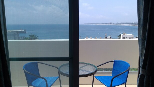 Varanda com duas cadeiras azuis e uma mesa pequena, com vista para a costa, edifícios e o mar.