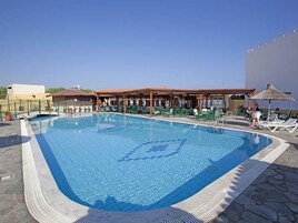 Piscina retangular com borda azulejada, rodeada por espreguiçadeiras e uma área de estar coberta.