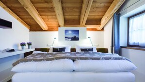 Quarto com teto de madeira, cama com roupa de cama branca, mesa de cabeceira com abajur e janela com cortinas.