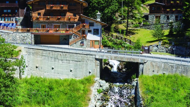 Um chalé de madeira com varanda, uma ponte de pedra e um riacho.