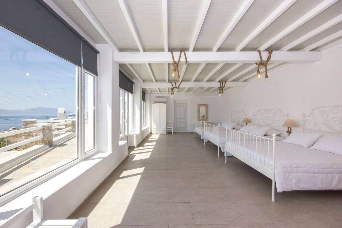 Quarto amplo e luminoso, com grandes janelas, uma cama longa e branca, e vista para o mar.
