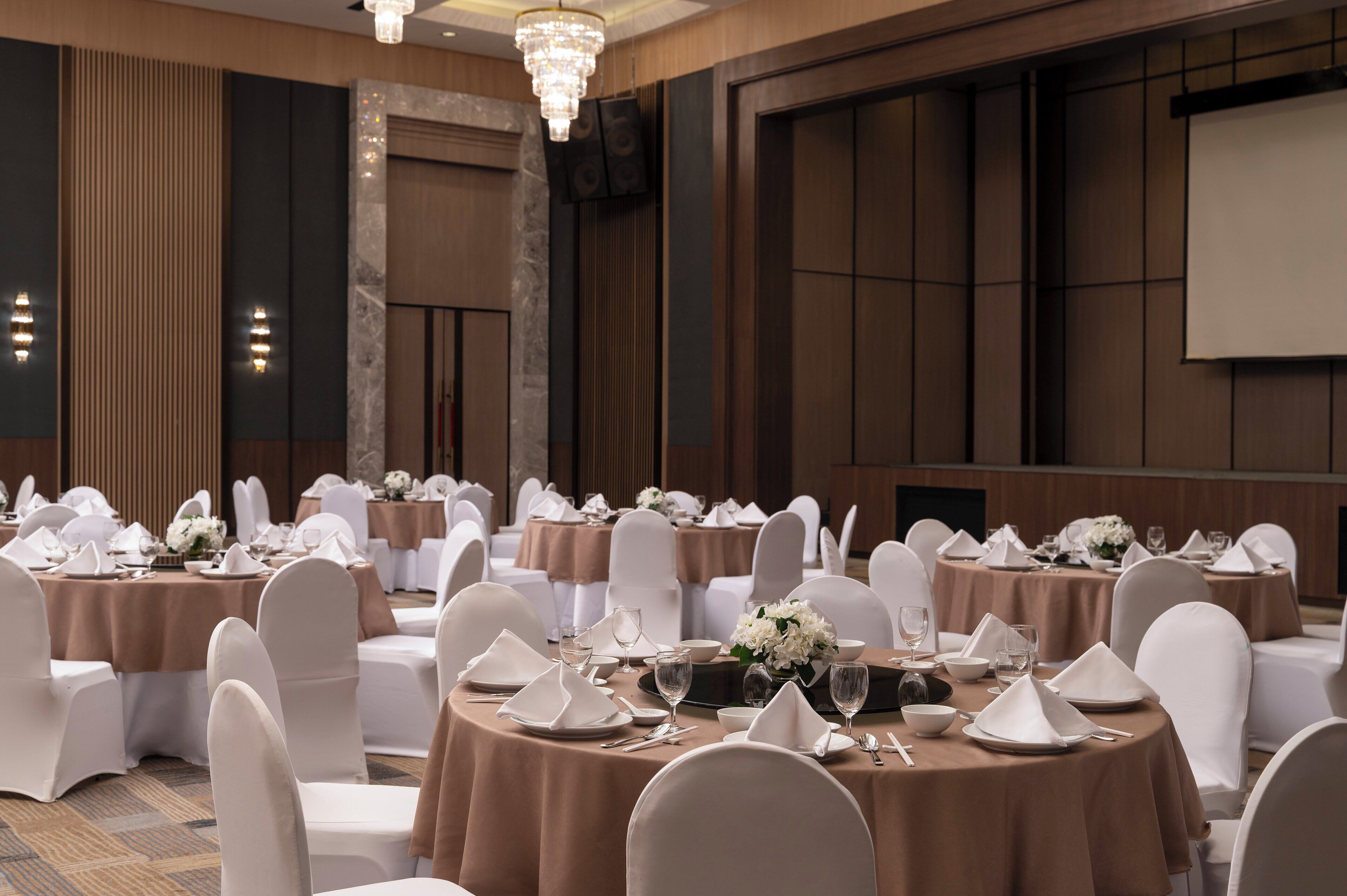 Uma sala de banquetes com mesas redondas dispostas para um evento formal, com cadeiras brancas e louças elegantes.