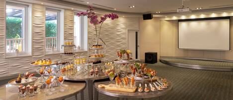 Uma sala de conferências com uma mesa de buffet exibindo diversos doces e petiscos, uma tela de projetor e um palestrante.