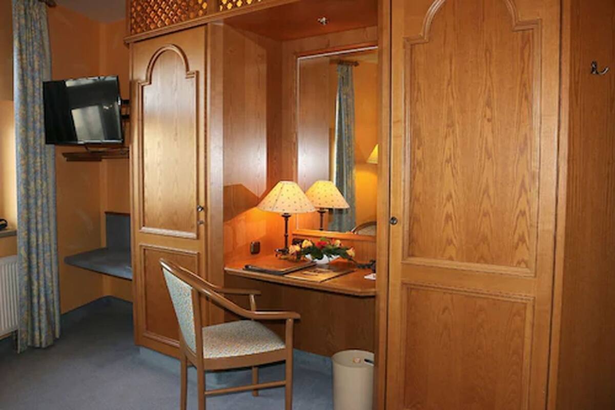 Quarto de hotel pequeno com uma escrivaninha de madeira, duas luminárias, uma cadeira, televisão e um espelho.
