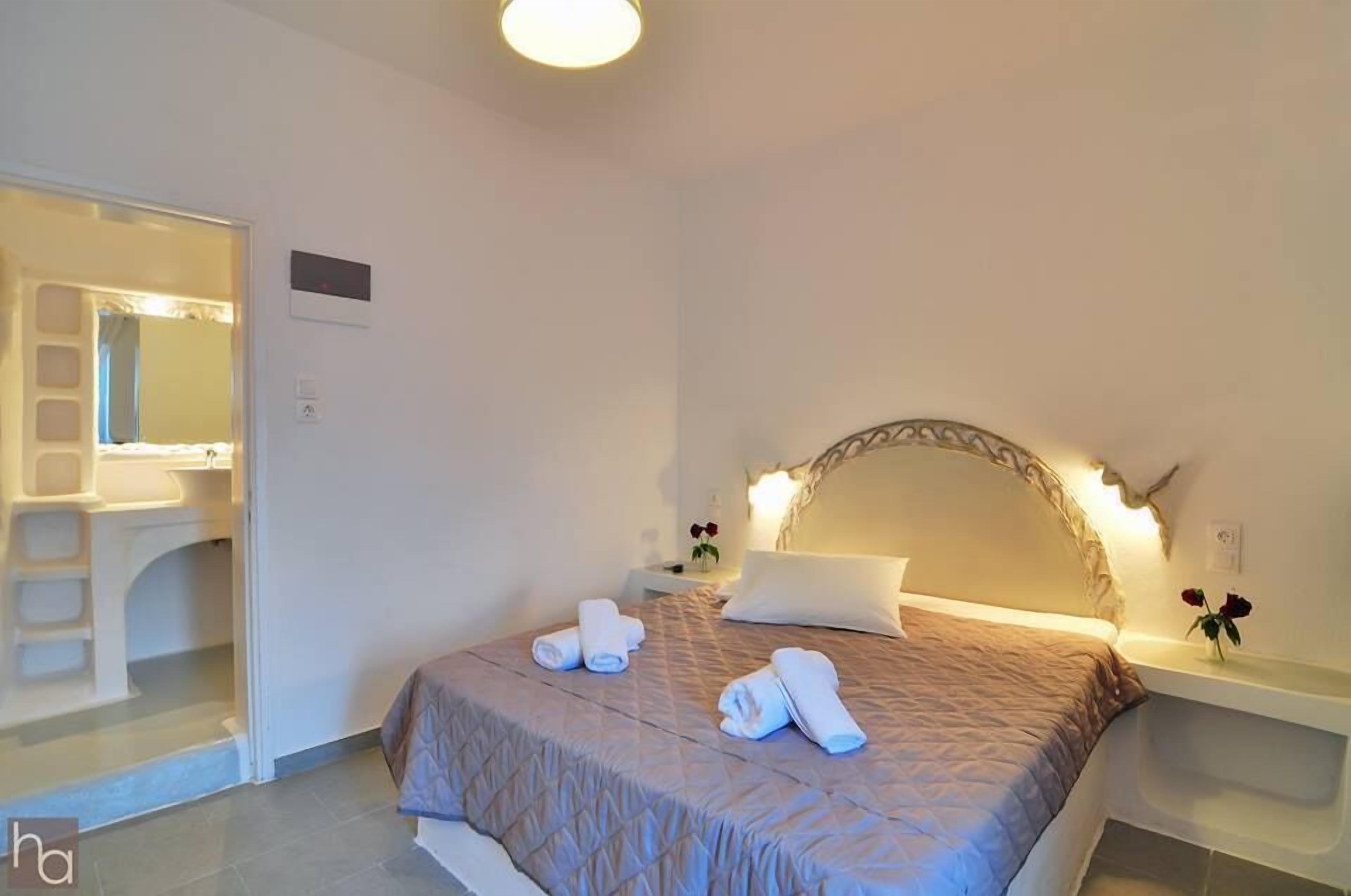 Cama bem arrumada com cabeceira, mesa de cabeceira com um vaso de flores e um banheiro com espelho e pia à vista.