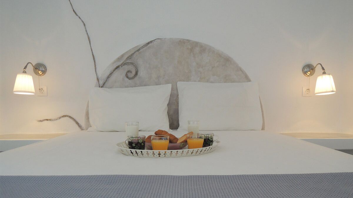Cama com cabeceira branca, duas luminárias na parede e bandeja com itens de café da manhã.