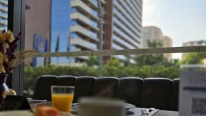 Uma mesa de jantar com uma chávena, um copo de sumo de laranja e um prato com comida. Há uma janela com vista para edifícios altos e vegetação.