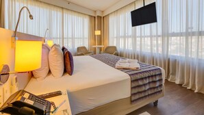 Quarto de hotel com cama, luminárias laterais, televisão, escrivaninha e cadeira.
