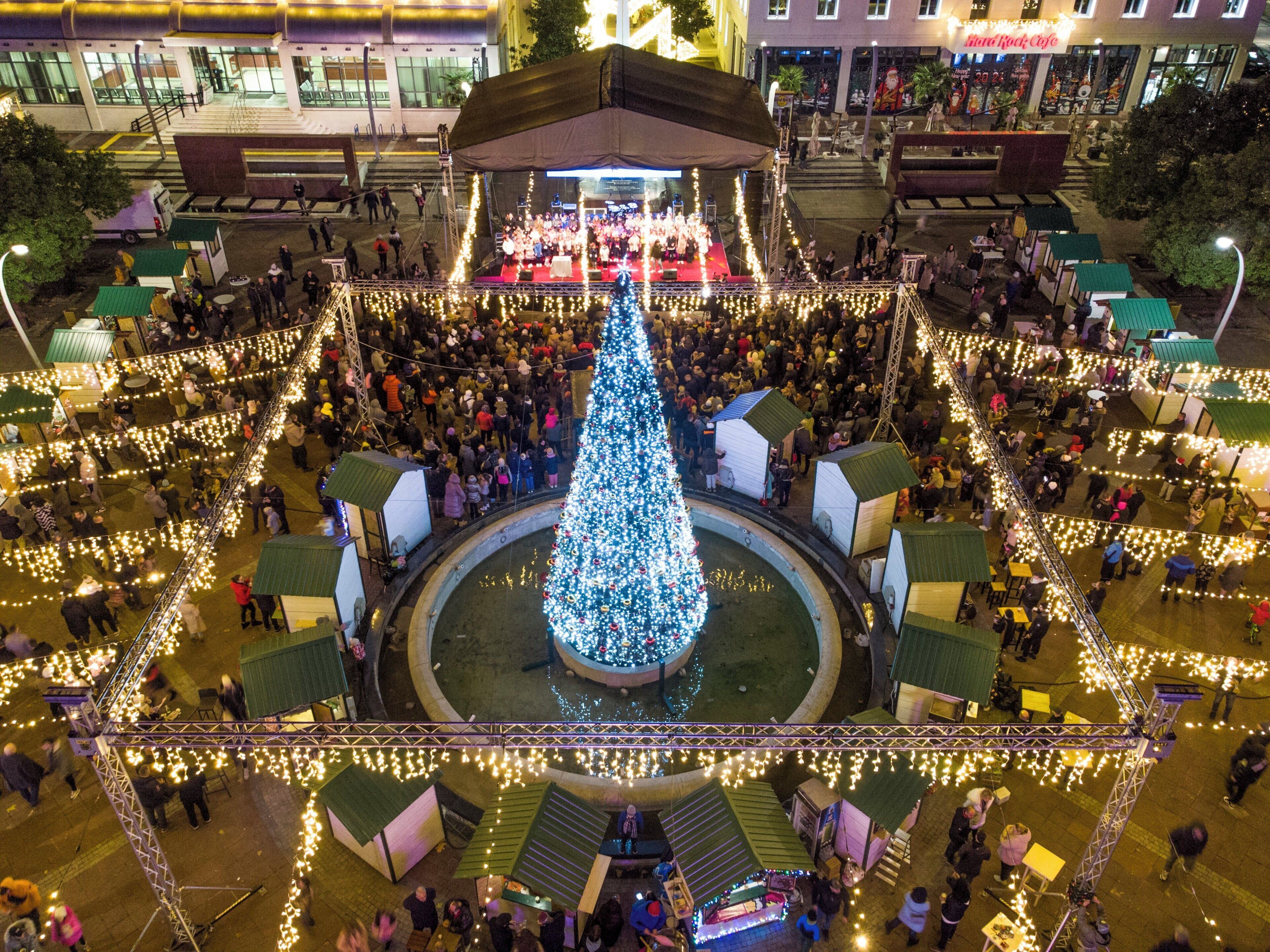 Uma árvore de Natal imensa numa praça pública, rodeada por pessoas e luzes festivas.