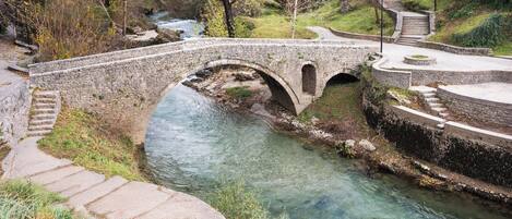 Uma ponte de pedra sobre um riacho límpido, rodeada por árvores e um caminho de pedra.