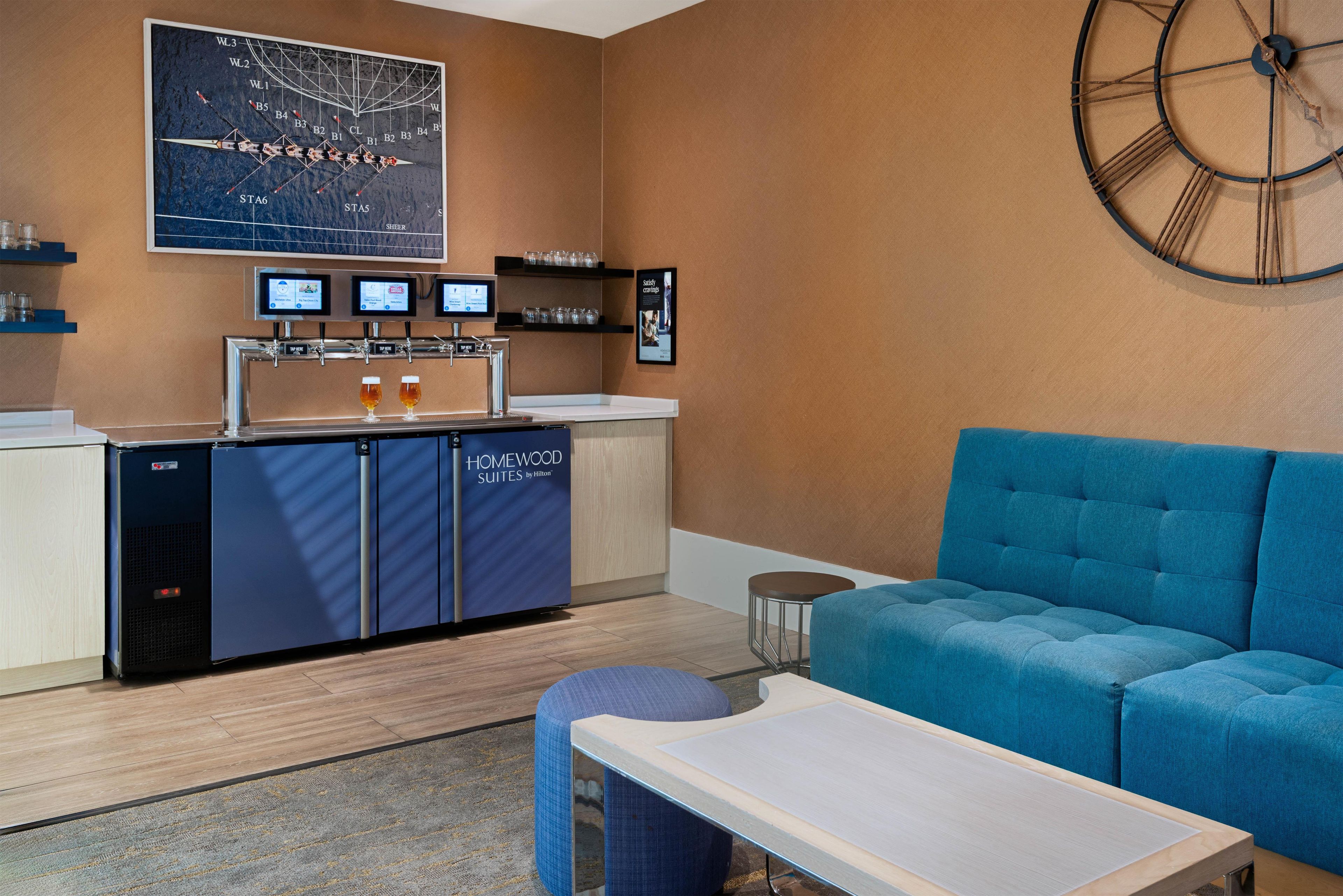 Quarto de hotel moderno com área de estar, minibar, máquina de café e um relógio de parede grande.