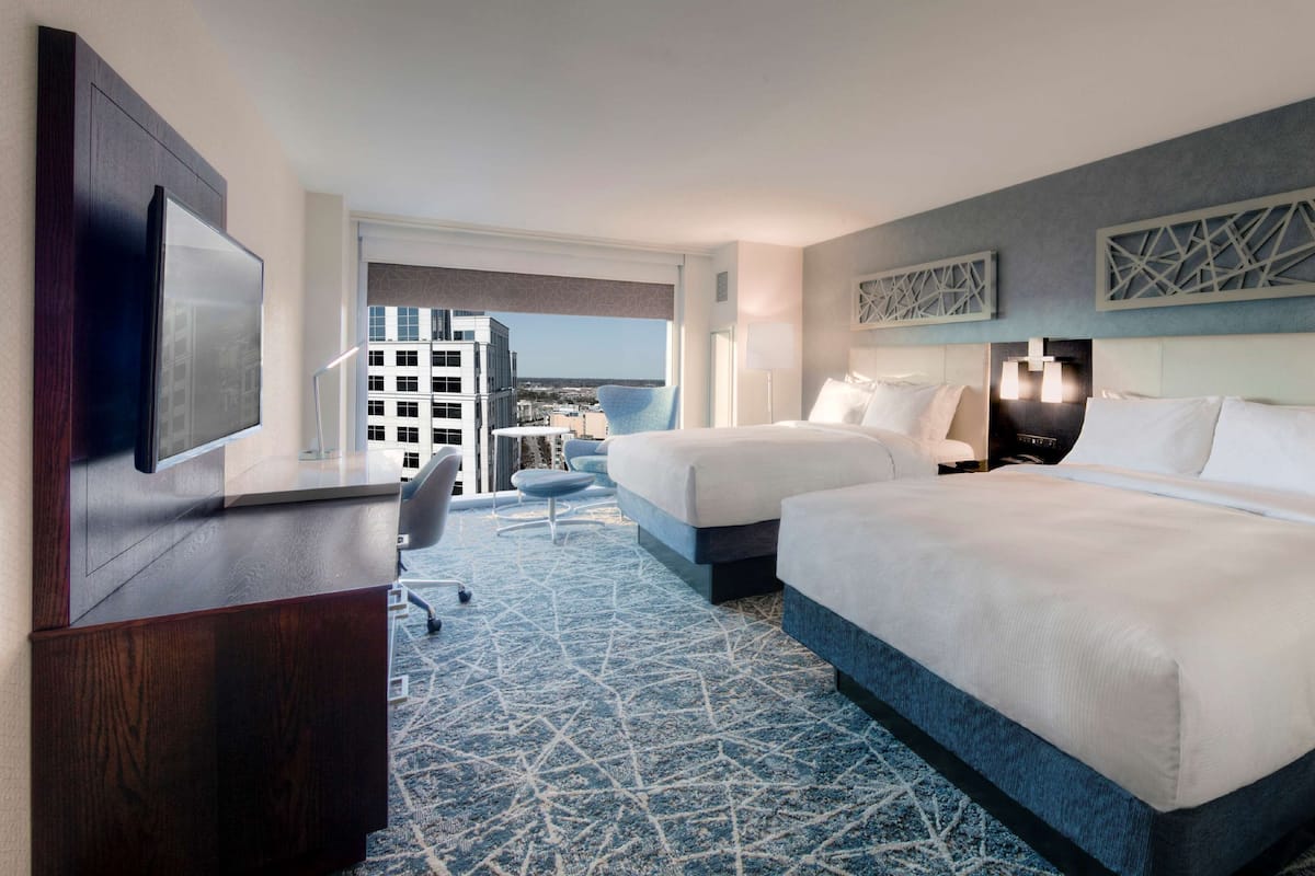 Quarto de hotel com duas camas, uma escrivaninha e televisão. Vê-se a cidade pela janela.