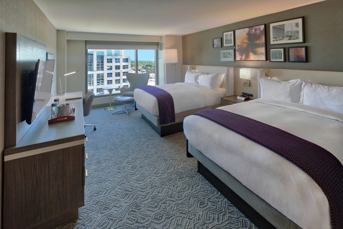 Quarto de hotel com duas camas, uma escrivaninha com televisão de tela plana, uma cadeira e uma janela grande com vista para a cidade.