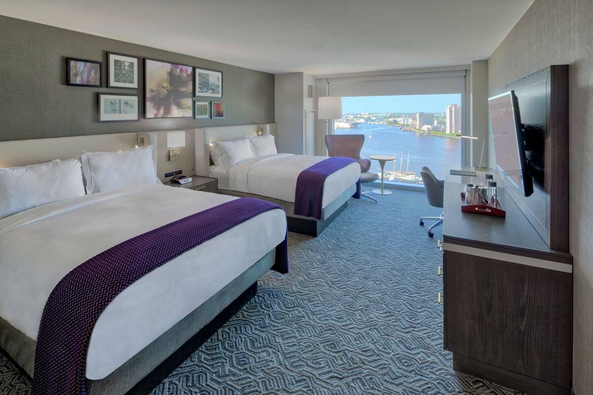 Quarto de hotel com duas camas, uma escrivaninha, uma cadeira, televisão e uma janela grande com vista para a cidade e para a água.