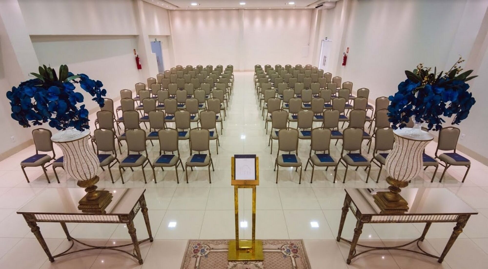 Uma sala de conferências com fileiras de cadeiras, dois vasos decorativos grandes e um pódio no centro.